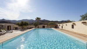Uma piscina com água cristalina, rodeada por espreguiçadeiras e plantas em vasos, com montanhas ao fundo.