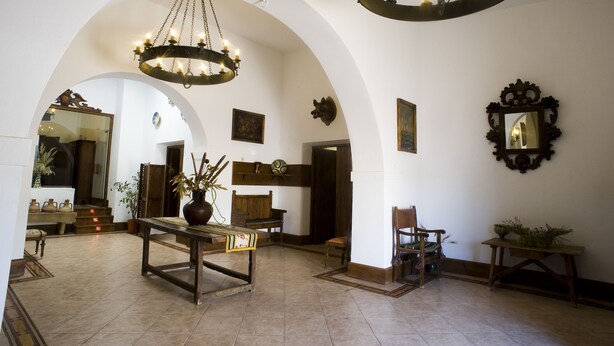 Um quarto amplo com teto alto abobadado, um banco de madeira, uma mesa com um vaso e um espelho na parede.