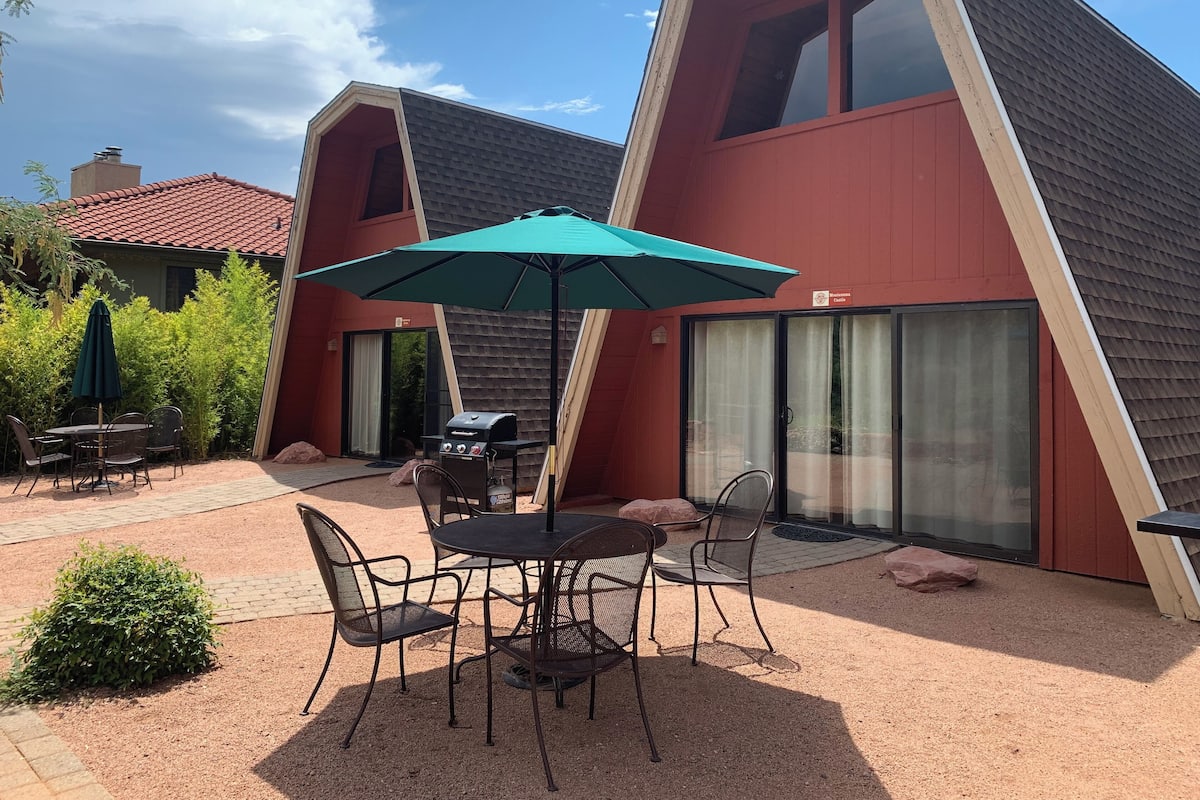 Um pátio externo com mesa, cadeiras, churrasqueira e um grande guarda-sol.