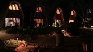 Fileira de cabanas pequenas e de formas originais, com grandes janelas, rodeadas por um jardim com plantas em vasos e uma pequena mesa.