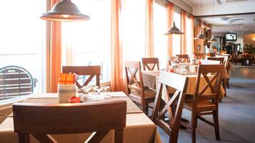 Um restaurante com mesas e cadeiras de madeira, iluminação pendente e vista para a rua através de grandes janelas.