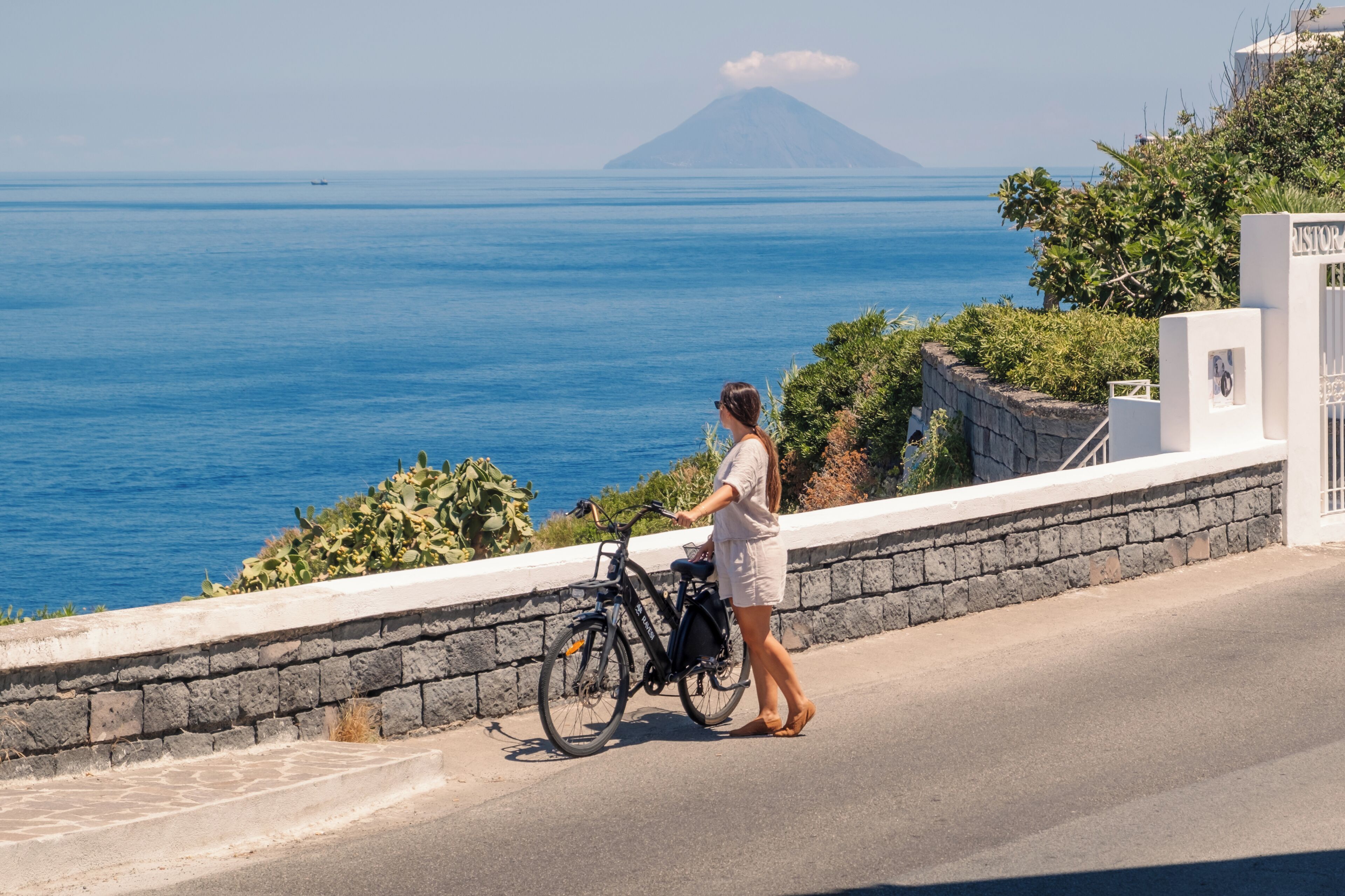 Uma pessoa ao lado de uma bicicleta numa estrada, com um muro de pedra e vegetação, com vista para um corpo de água e uma montanha ao longe.