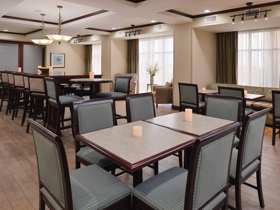 Uma sala de jantar de hotel com várias mesas e cadeiras, um candelabro e janelas amplas com cortinas.