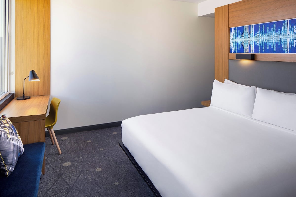 Quarto de hotel com uma cama, uma escrivaninha com luminária, uma cadeira e uma televisão fixada na parede.