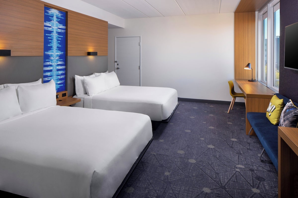 Quarto de hotel com duas camas, uma escrivaninha, uma cadeira, uma televisão e um painel luminoso vertical na parede.