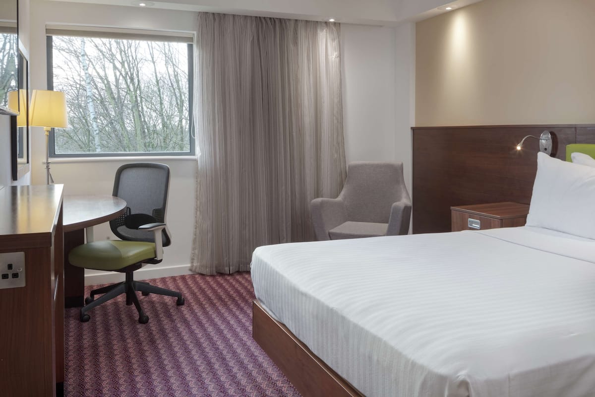 Quarto de hotel com cama, escrivaninha com cadeira, mesa de cabeceira e janela com cortinas.