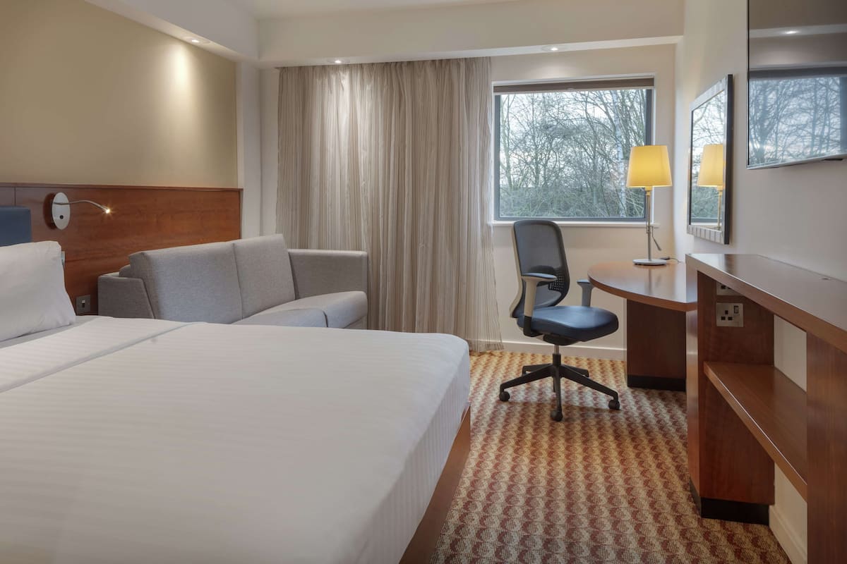 Quarto de hotel com uma cama grande, uma escrivaninha com cadeira, uma luminária e uma janela com cortinas.