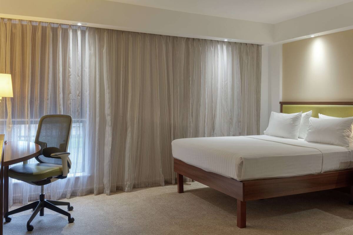 Quarto de hotel com uma cama, uma escrivaninha com cadeira e uma janela com cortinas.