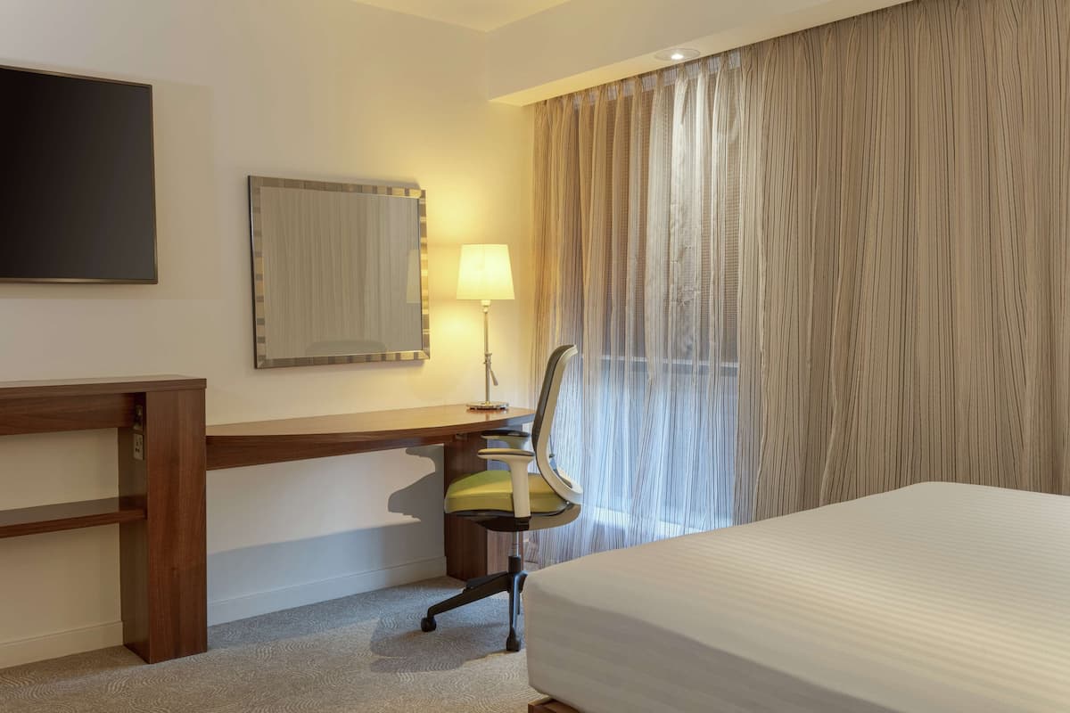 Quarto de hotel com uma cama, uma escrivaninha, uma cadeira, um abajur e uma janela grande com cortinas.