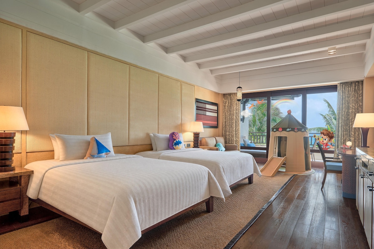Quarto de hotel com duas camas, uma escrivaninha, uma cadeira, espaço para brincar e vista para o exterior.
