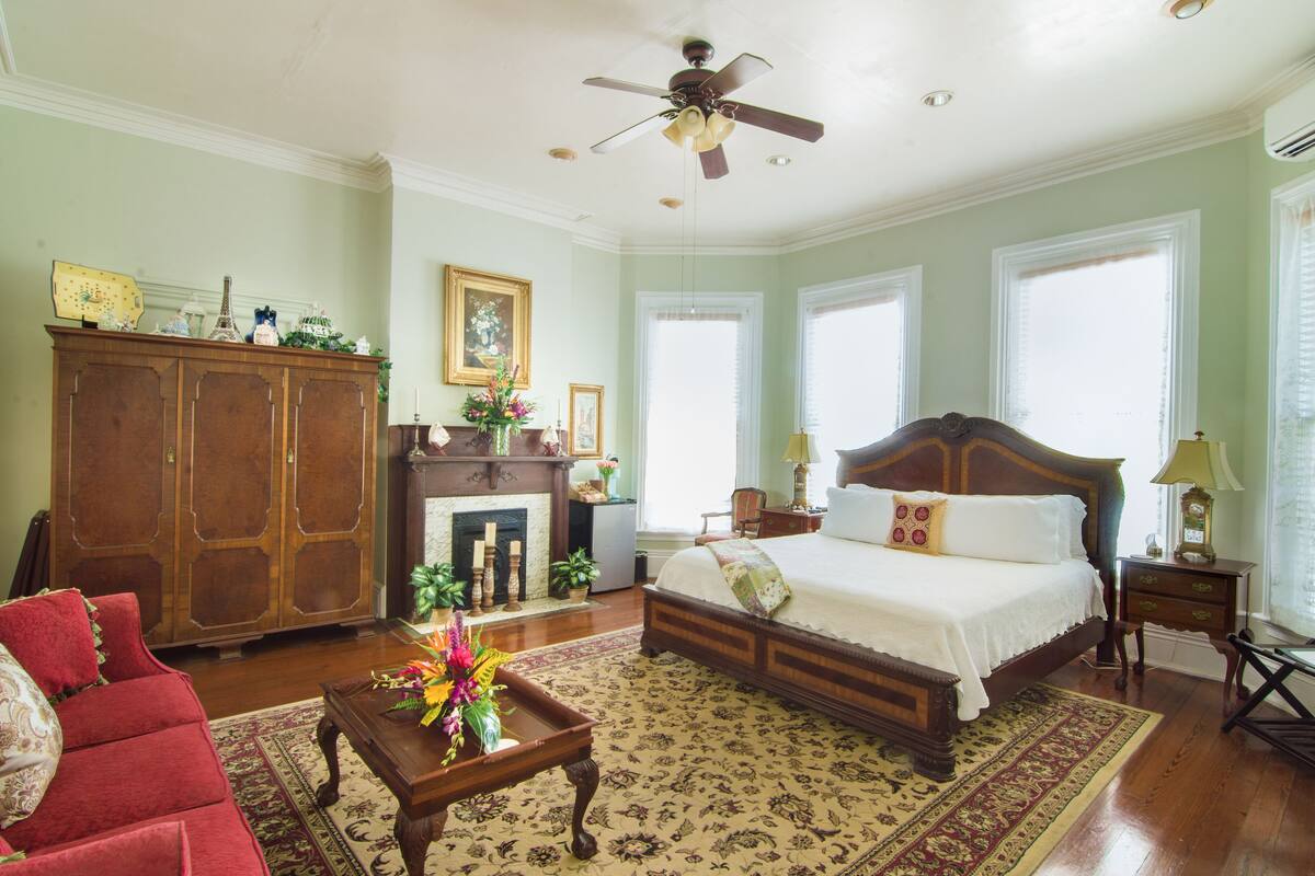 Quarto com uma cama grande, cômoda de madeira, poltrona vermelha, mesa de centro de madeira e ventilador de teto.
