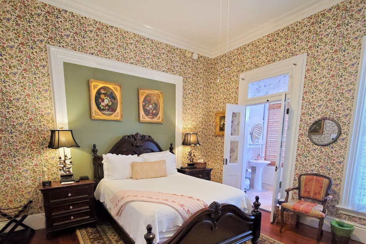 Quarto com cama grande, piso de madeira, papel de parede floral, ventilador de teto e porta que dá para outro cômodo.