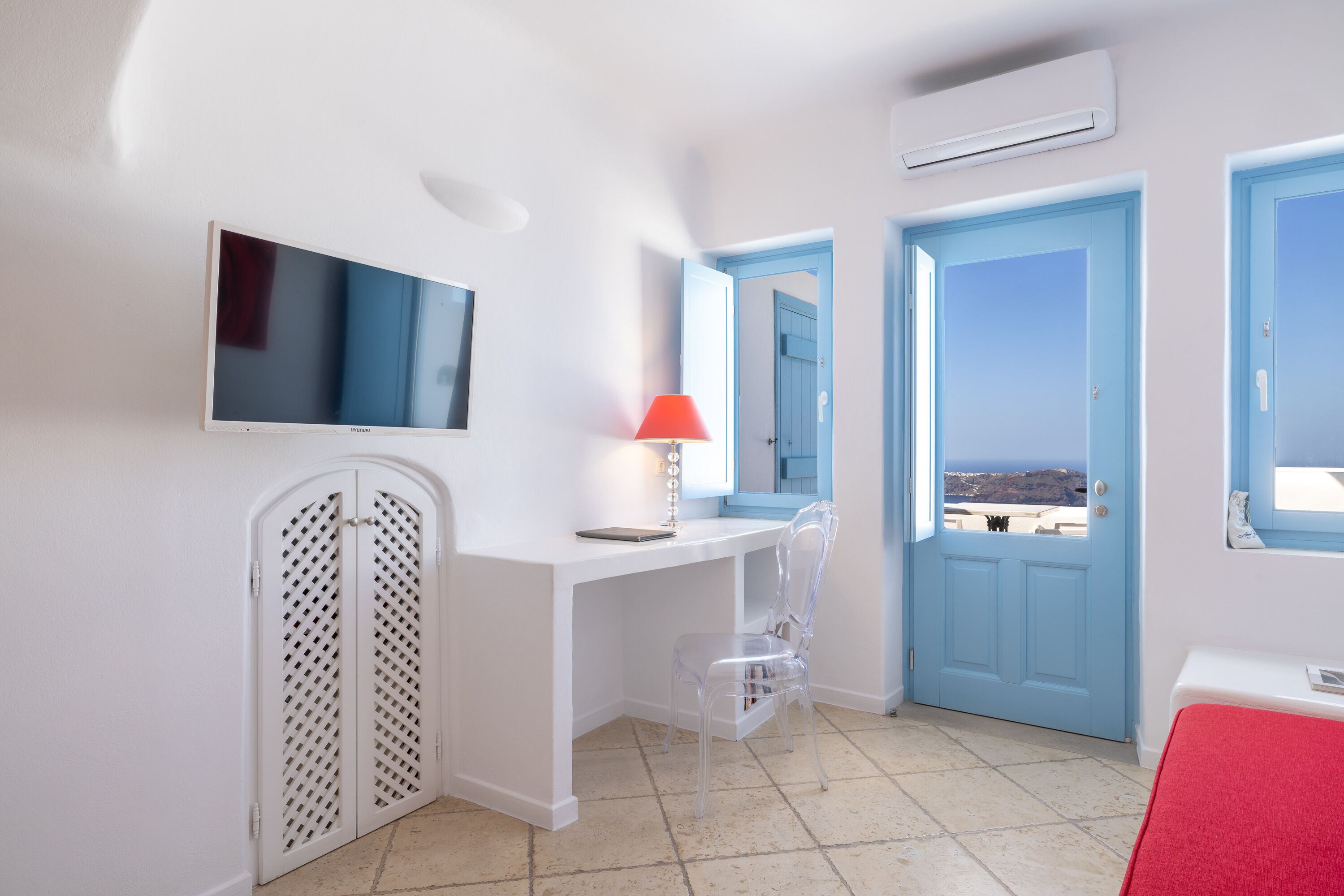 Quarto com uma escrivaninha branca, luminária vermelha, TV e porta que dá para uma varanda com vista.