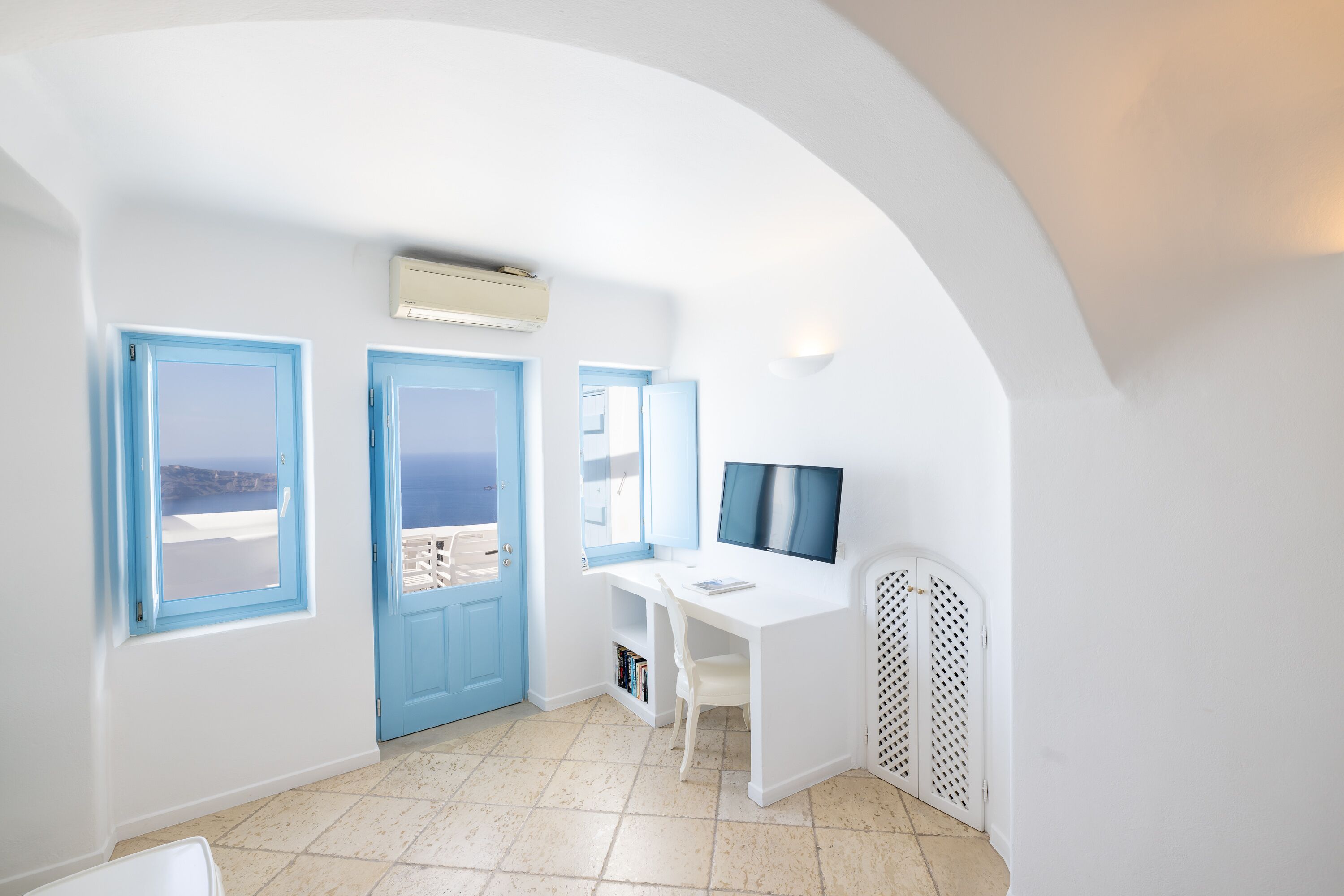 Quarto com uma escrivaninha branca, uma cadeira, uma televisão e vista para o mar através de uma porta.
