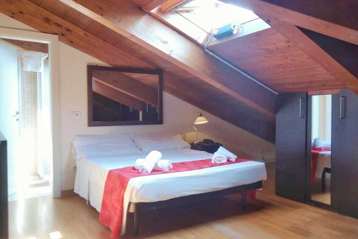 Um quarto com teto inclinado, uma cama com manta vermelha, abajur e espelho na parede.