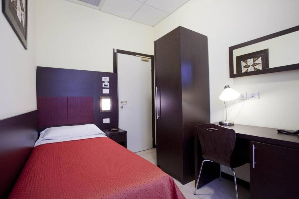 Quarto de hotel com cama, escrivaninha, guarda-roupa e mesa de cabeceira.