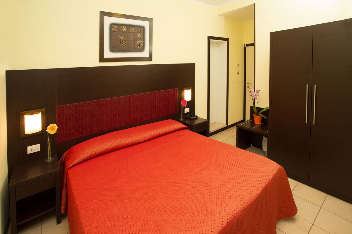 Quarto de hotel com uma cama grande, mesa de cabeceira com abajur, espelho e guarda-roupa.