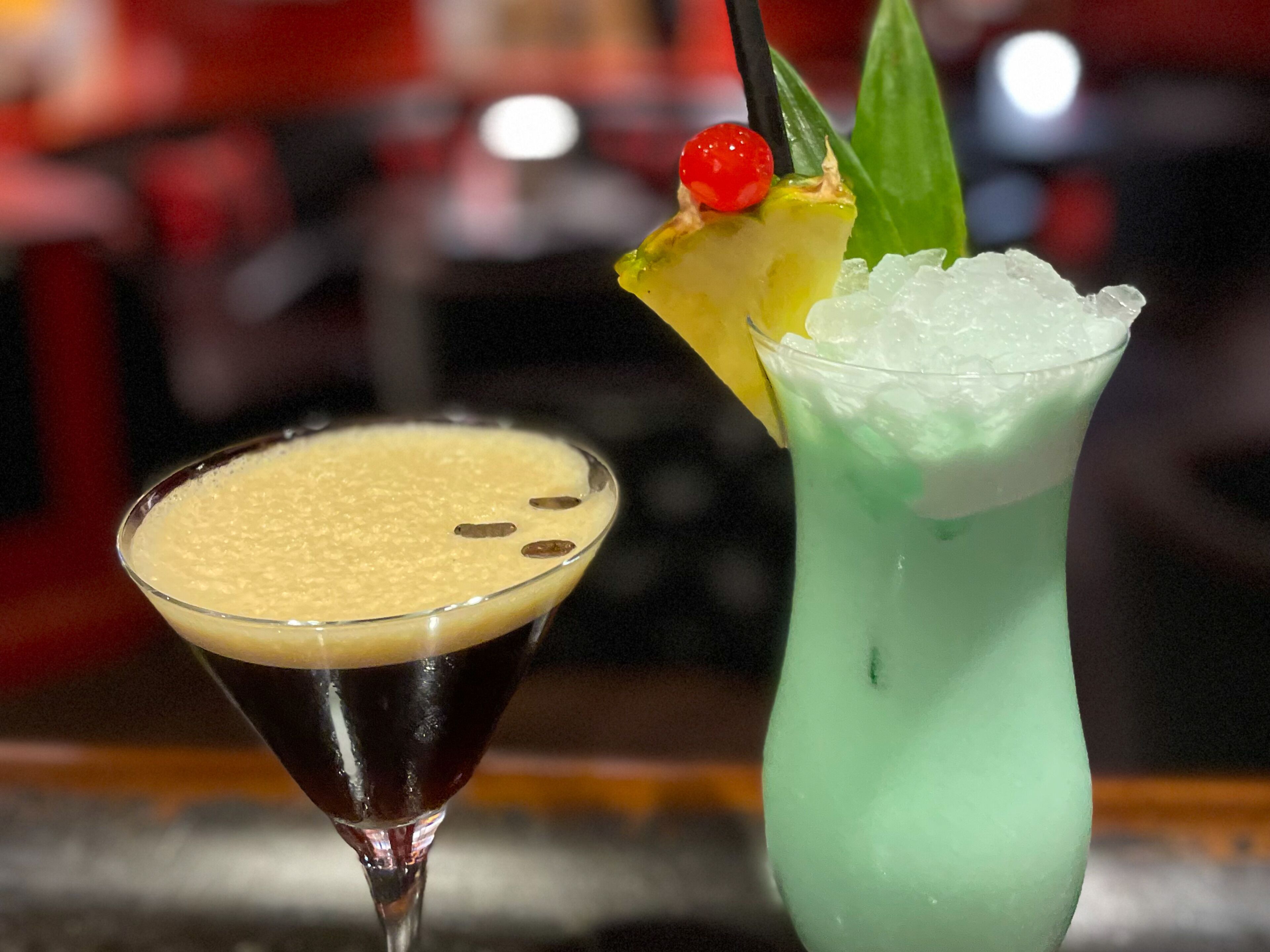 Dois cocktails em copos de martini, um com guarnição amarela e outro com guarnição verde, dispostos sobre um balcão de bar.