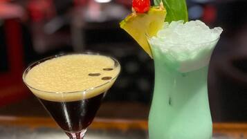 Dois cocktails em copos de martini, um com guarnição amarela e outro com guarnição verde, dispostos sobre um balcão de bar.