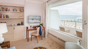 Quarto de hotel com escrivaninha, uma cadeira, mesa de centro com fruta, estante de livros, televisão e uma ampla janela com vista para o mar e um cais.