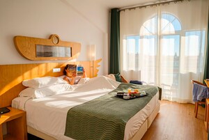 Quarto de hotel com cama, bandeja de café da manhã, janela com cortinas e relógio fixado na parede.