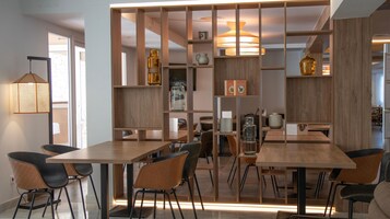Uma área de refeições moderna com mesas e cadeiras de madeira, uma divisória decorativa com prateleiras e uma luminária suspensa.