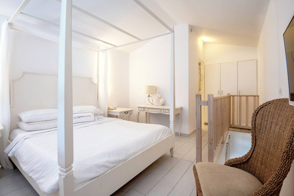 Uma cama de dossel com roupa de cama branca, uma mesa de cabeceira com abajur, uma cadeira de vime e uma escada de madeira.