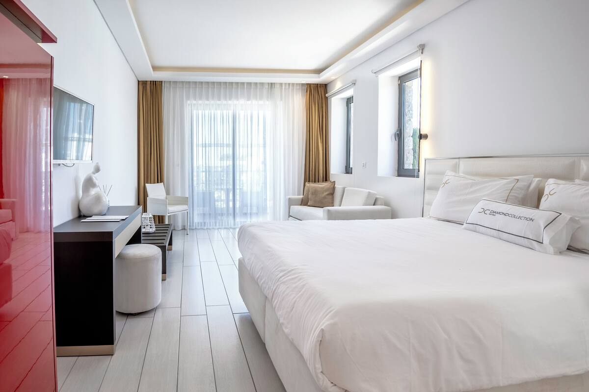 Quarto de hotel moderno com uma cama grande, uma escrivaninha com cadeira, uma TV e vista para o exterior através de amplas janelas.