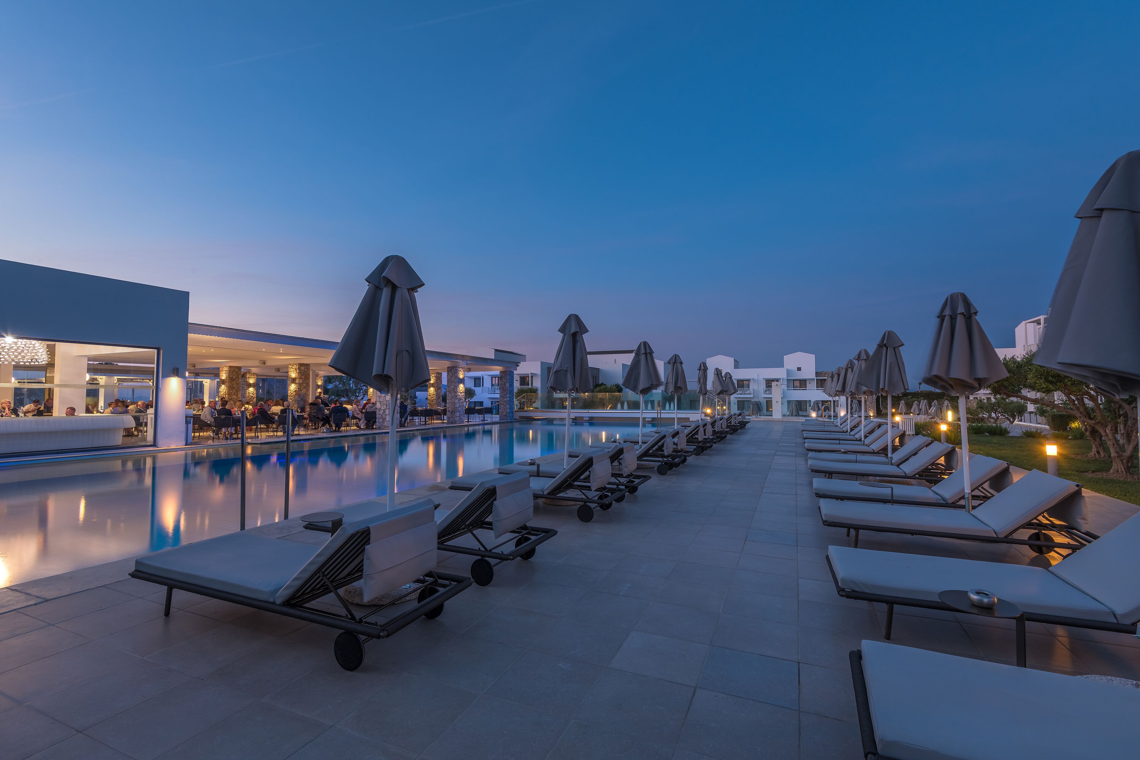 Uma área ao lado da piscina com espreguiçadeiras e guarda-sóis, uma piscina e um edifício com mesas externas.
