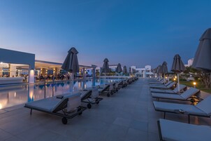 Uma área ao lado da piscina com espreguiçadeiras e guarda-sóis, uma piscina e um edifício com mesas externas.