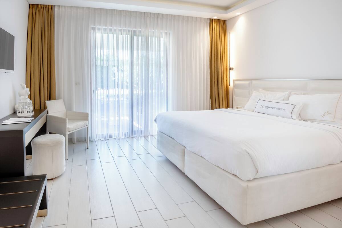 Quarto de hotel com uma cama grande, uma escrivaninha com cadeira e vista através de cortinas leves.