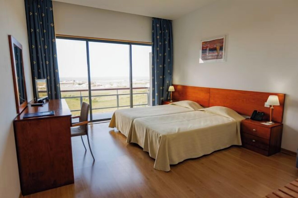 Quarto de hotel com duas camas, uma escrivaninha, uma cadeira, mesa de cabeceira e uma janela grande com cortinas.