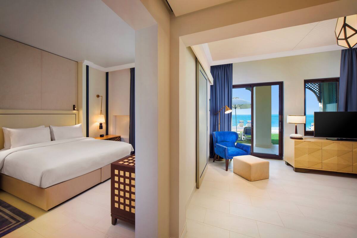 Quarto de hotel com uma cama grande, televisão, área de estar com uma poltrona e apoio para os pés, e vista para a praia.