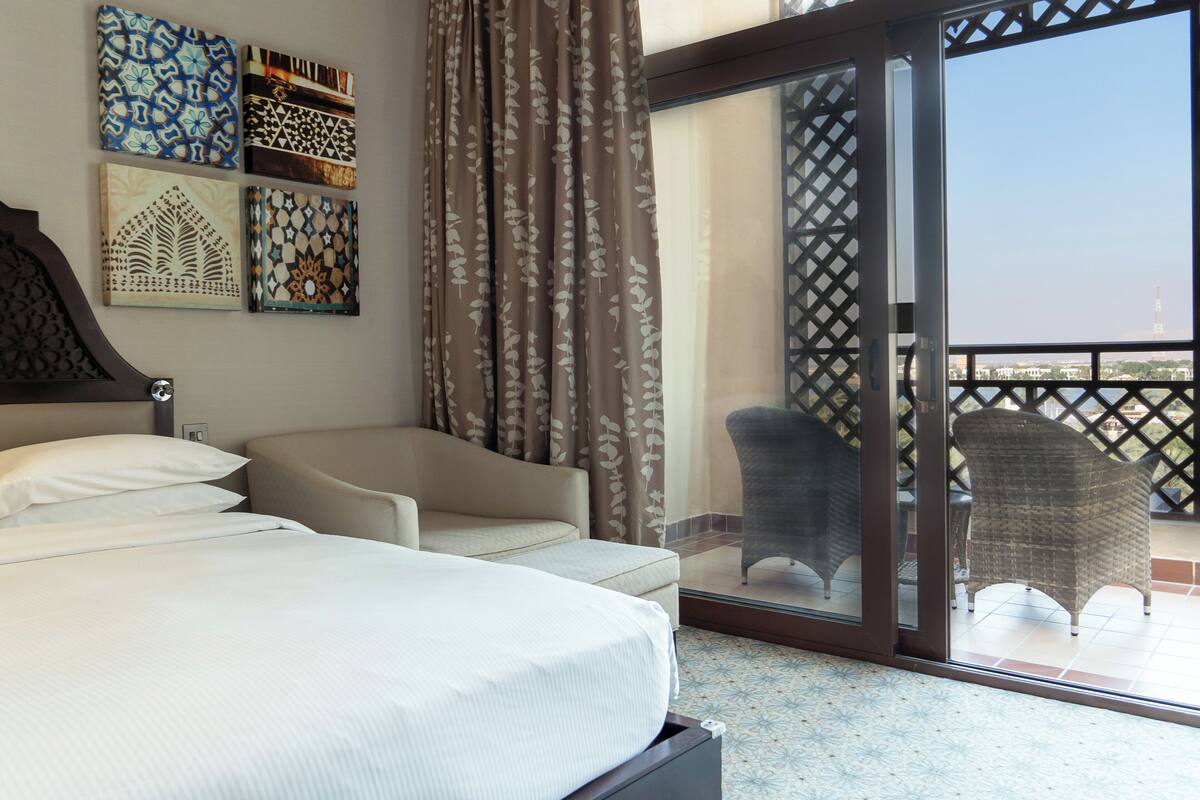 Quarto de hotel com cama, sofá, varanda com móveis de vime e arte decorativa na parede.