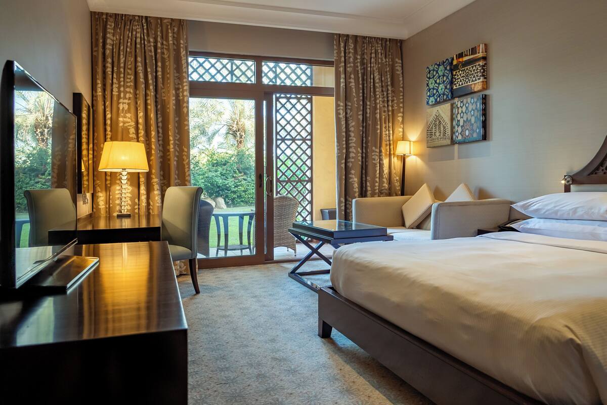 Quarto de hotel com cama, escrivaninha, cadeira e mesa. Há cortinas e uma janela com vista para vegetação.