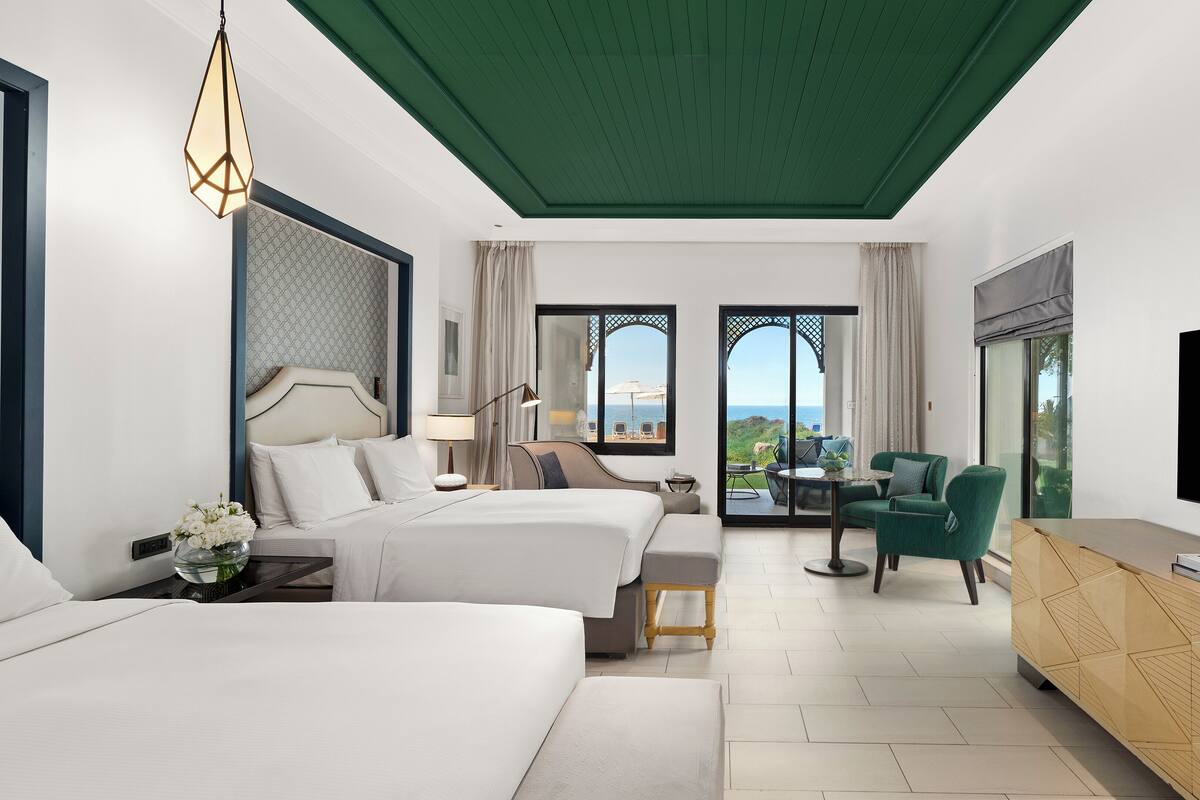Quarto de hotel moderno com duas camas, televisão, área de refeições com mesa e cadeiras, e vista para o mar.