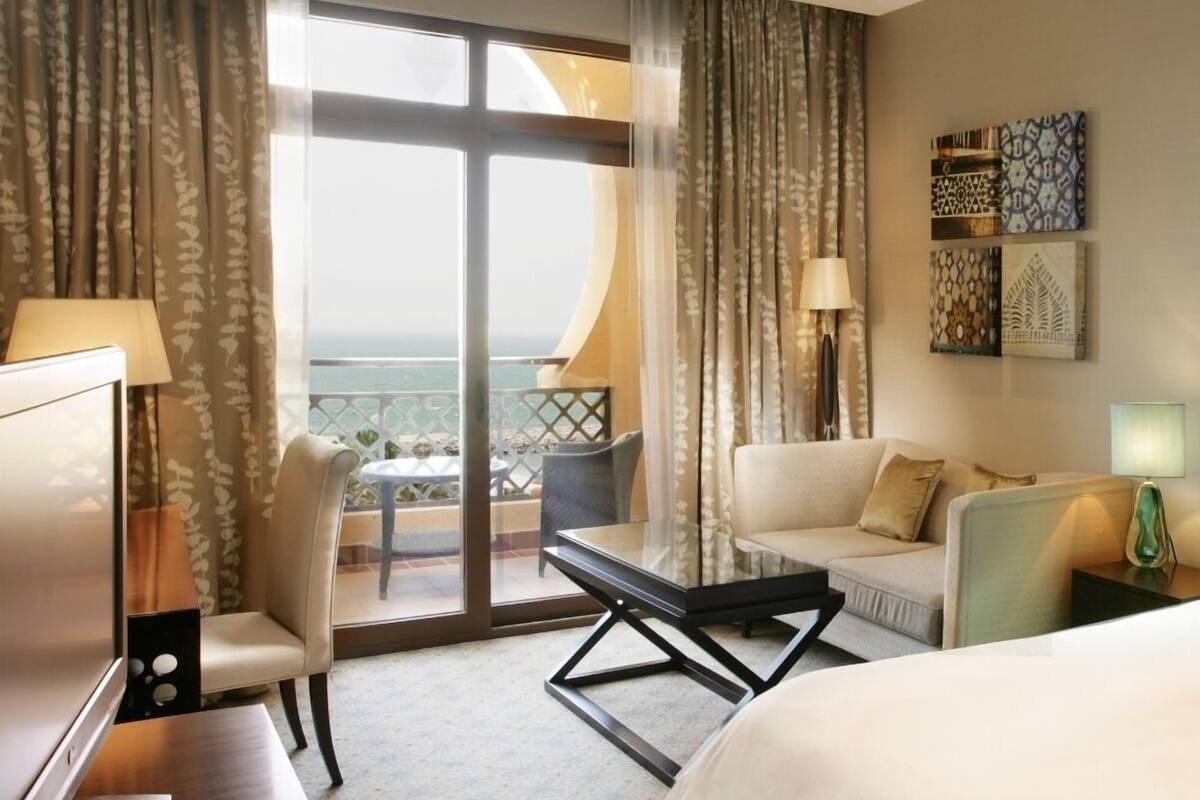 Quarto de hotel com cama, escrivaninha, poltrona, sofá, televisão e varanda vista para o mar.