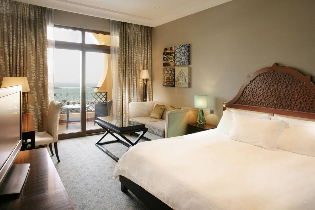 Quarto de hotel com cama, sofá, escrivaninha e vista para o mar através de grandes janelas.
