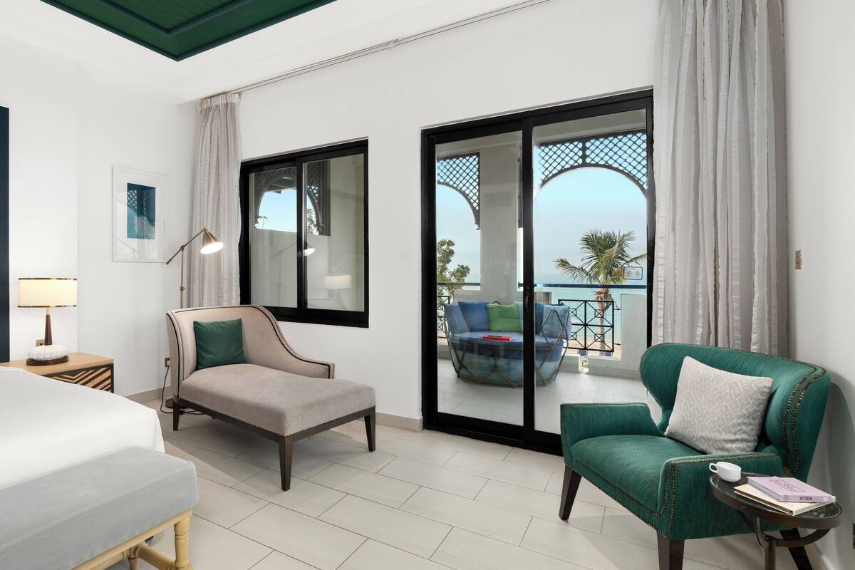 Quarto de hotel com cama, poltrona, sofá, varanda e vista para o mar, e uma mesa com um livro e uma xícara.