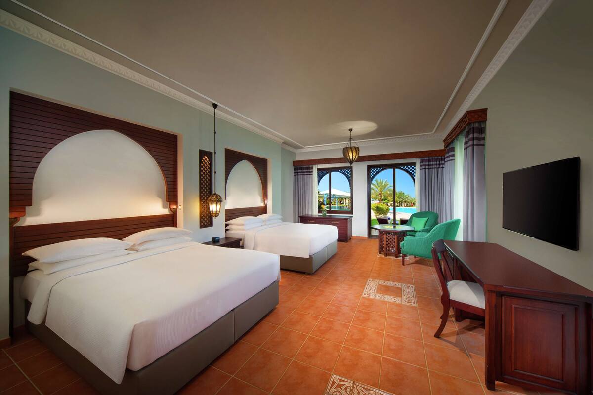 Quarto de hotel com duas camas, televisão, escrivaninha e vista para o exterior.