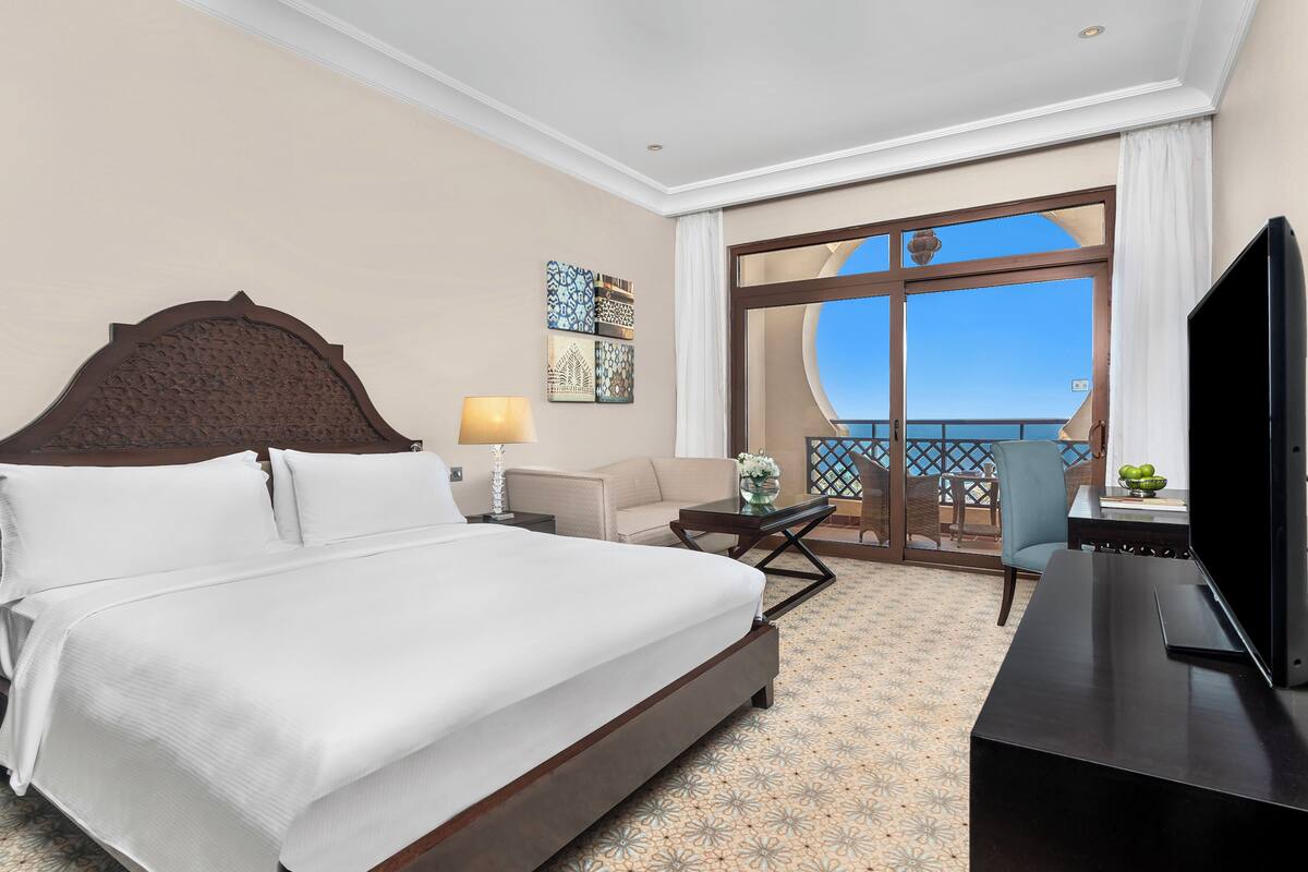 Quarto de hotel com uma cama grande, uma televisão, varanda com vista para o mar e uma pequena mesa com abajur.