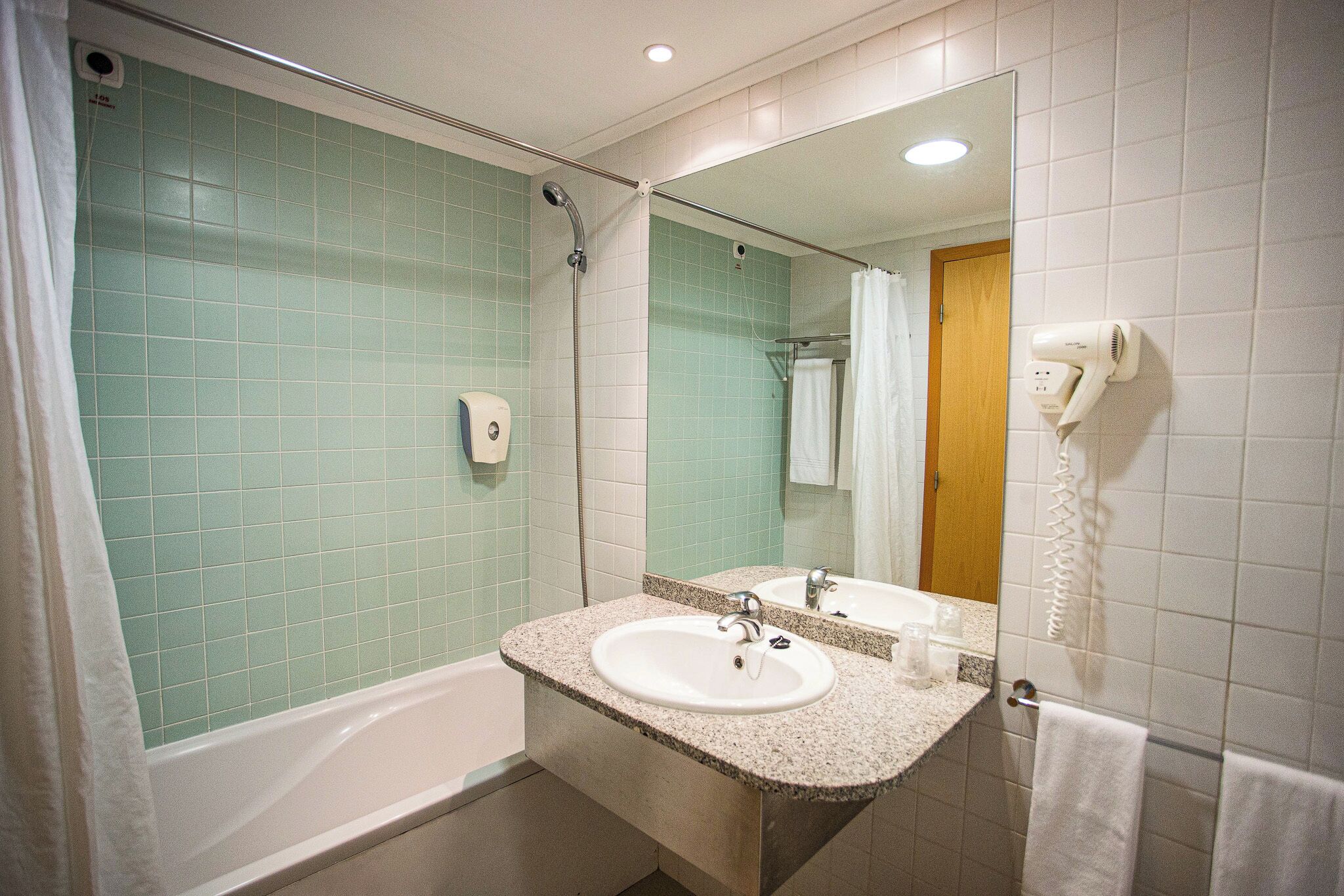 Uma casa de banho com banheira, pia, espelho e secador de cabelo fixado na parede.