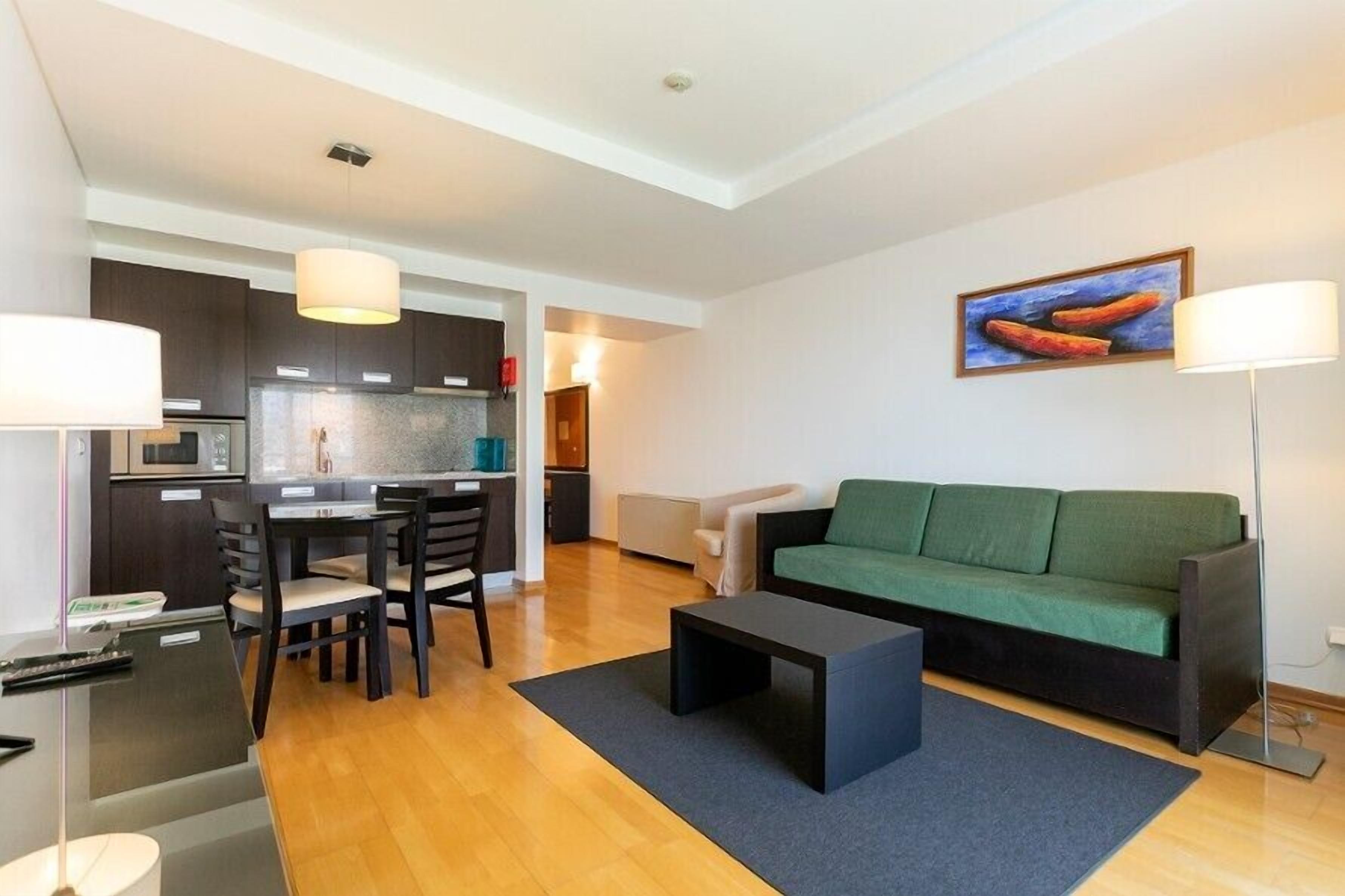 Uma sala de estar moderna com um sofá verde, uma mesa de jantar com cadeiras, uma mesa de centro e uma área de cozinha ao fundo.