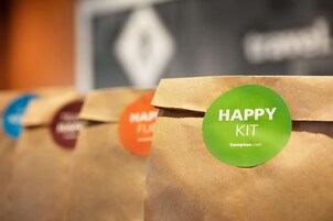 Uma fileira de sacos de papel castanhos com autocolantes verdes, um deles com a inscrição 'HAPPY KIT' e os outros com diferentes etiquetas.