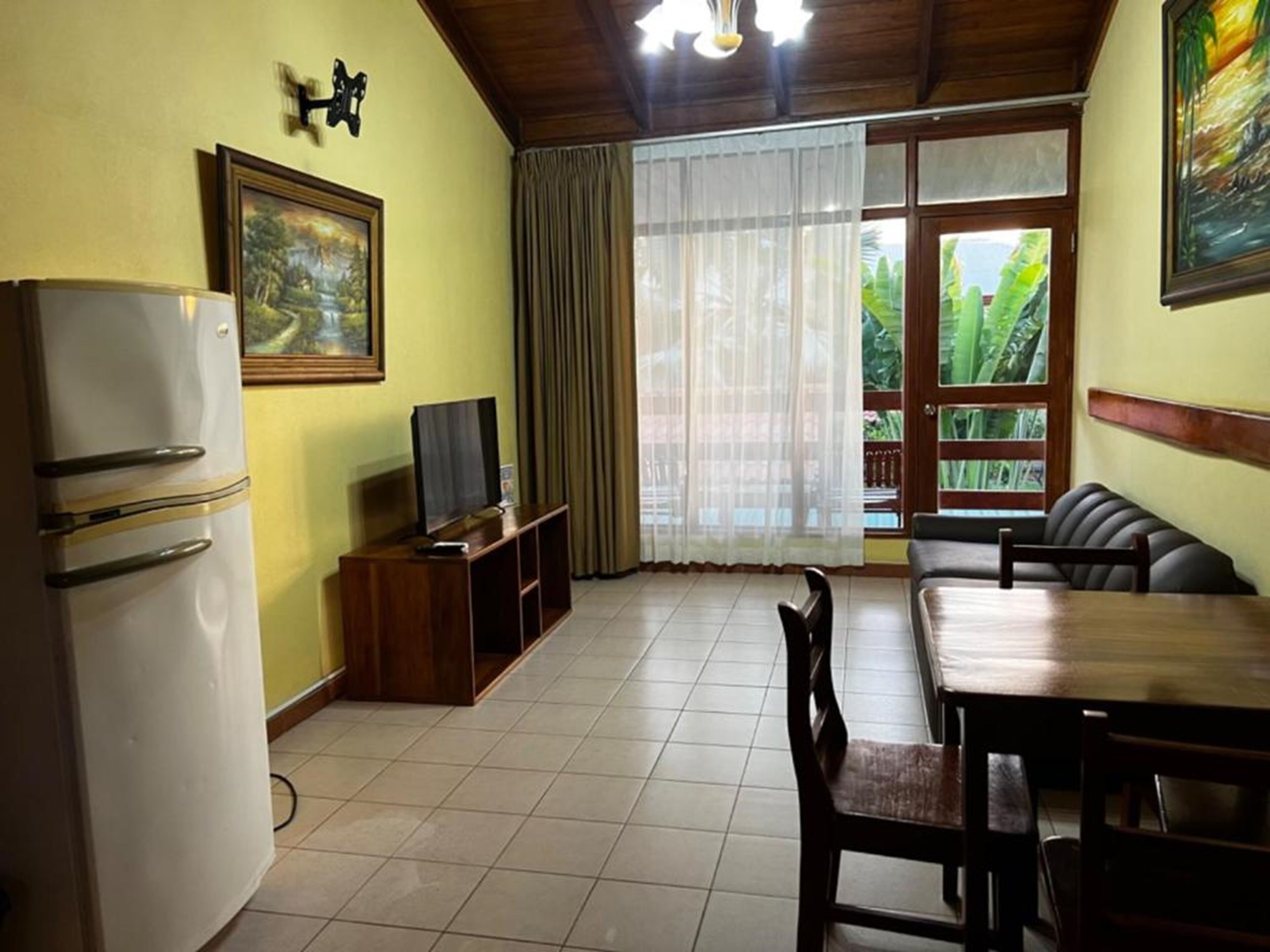 Um espaço de estar compacto com área de jantar, uma televisão sobre um suporte de madeira, um refrigerador e uma janela grande com cortinas.