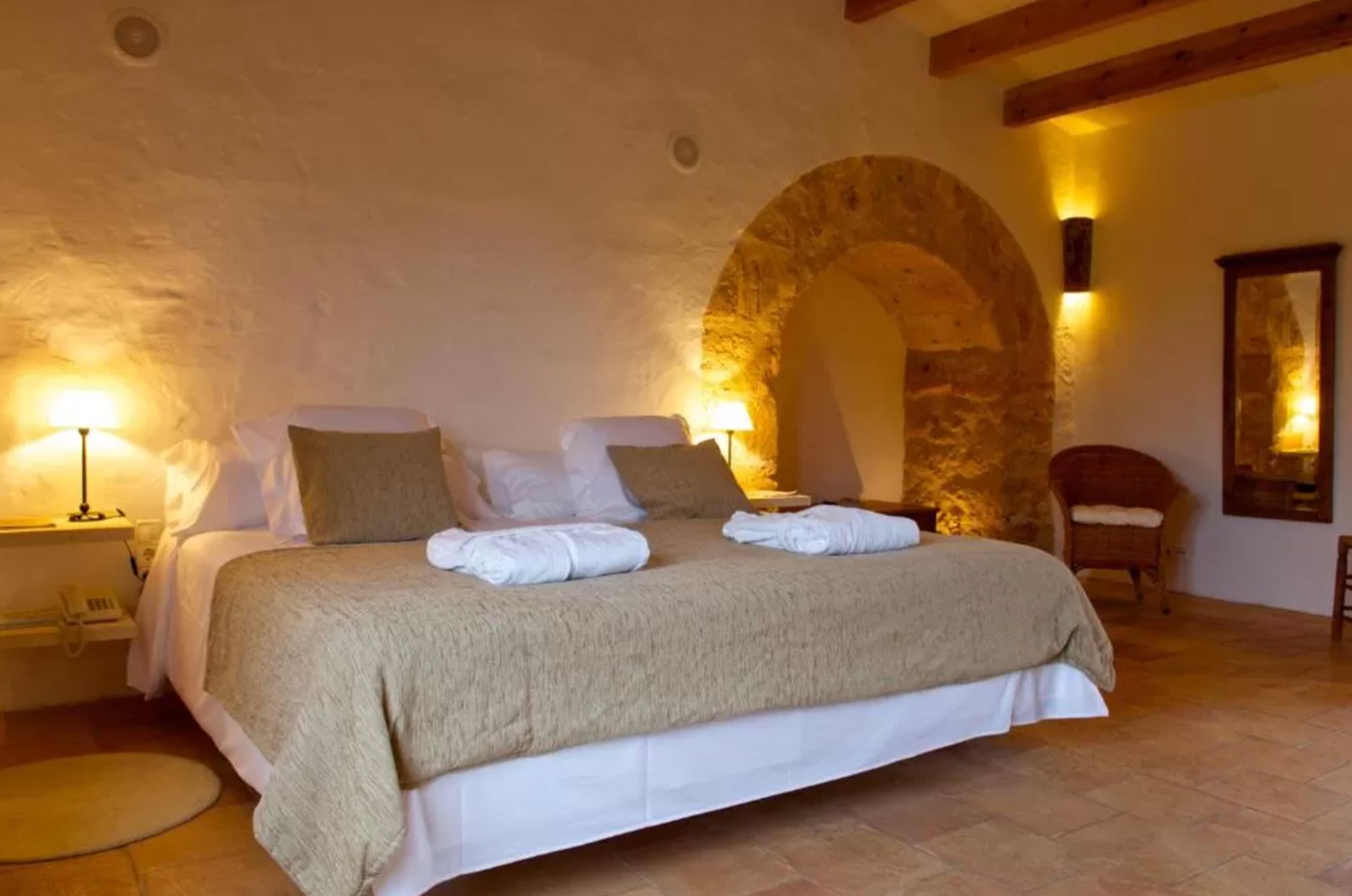 Quarto aconchegante com arco de pedra, cama com lençóis brancos, duas luminárias de cabeceira e uma cadeira de madeira.
