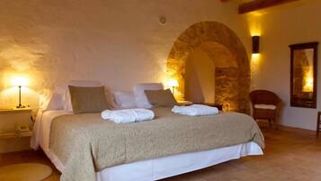 Quarto aconchegante com arco de pedra, cama com lençóis brancos, duas luminárias de cabeceira e uma cadeira de madeira.