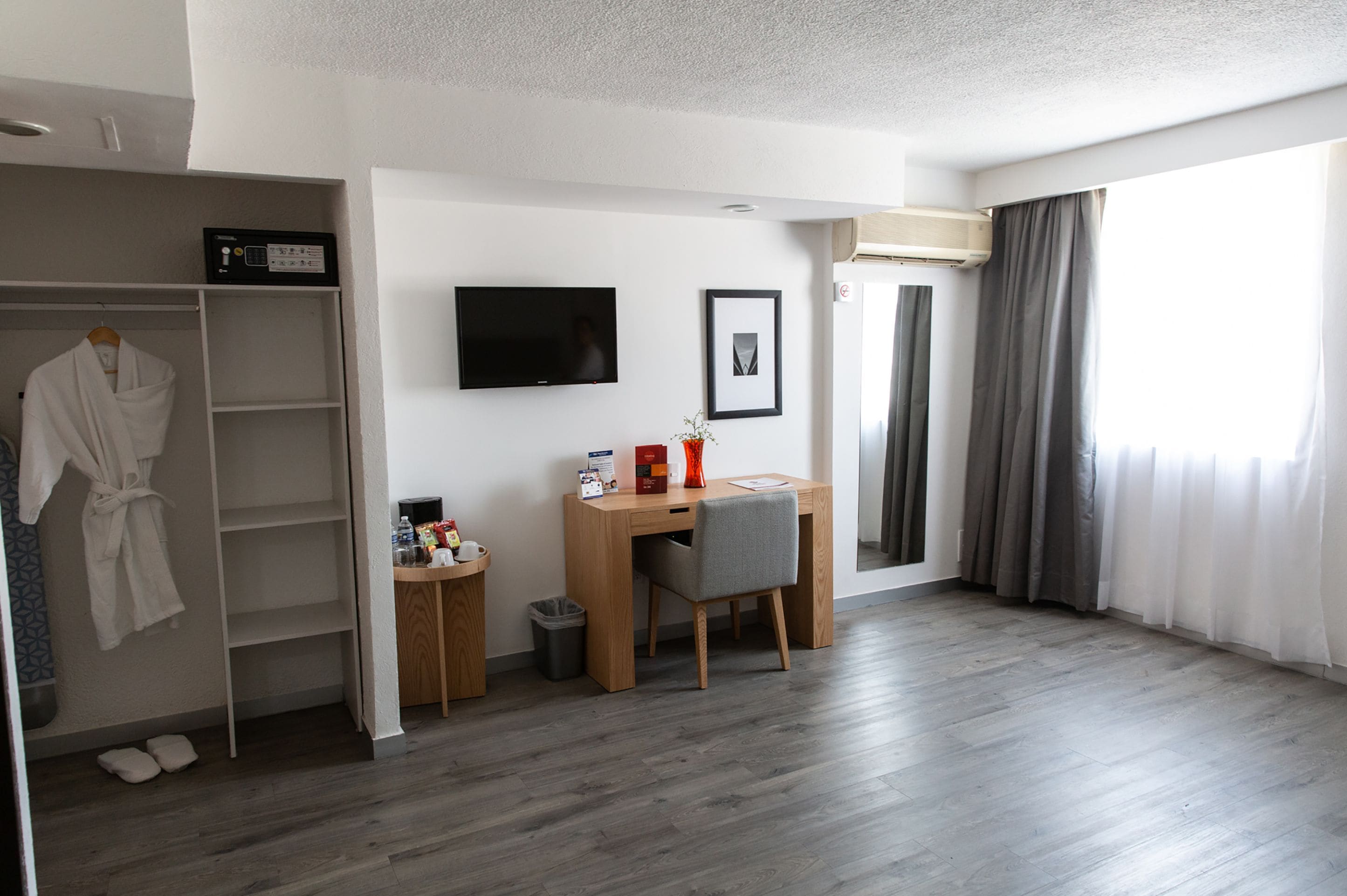 Quarto de hotel com escrivaninha de madeira, televisão, guarda-roupa e uma janela grande com cortinas.