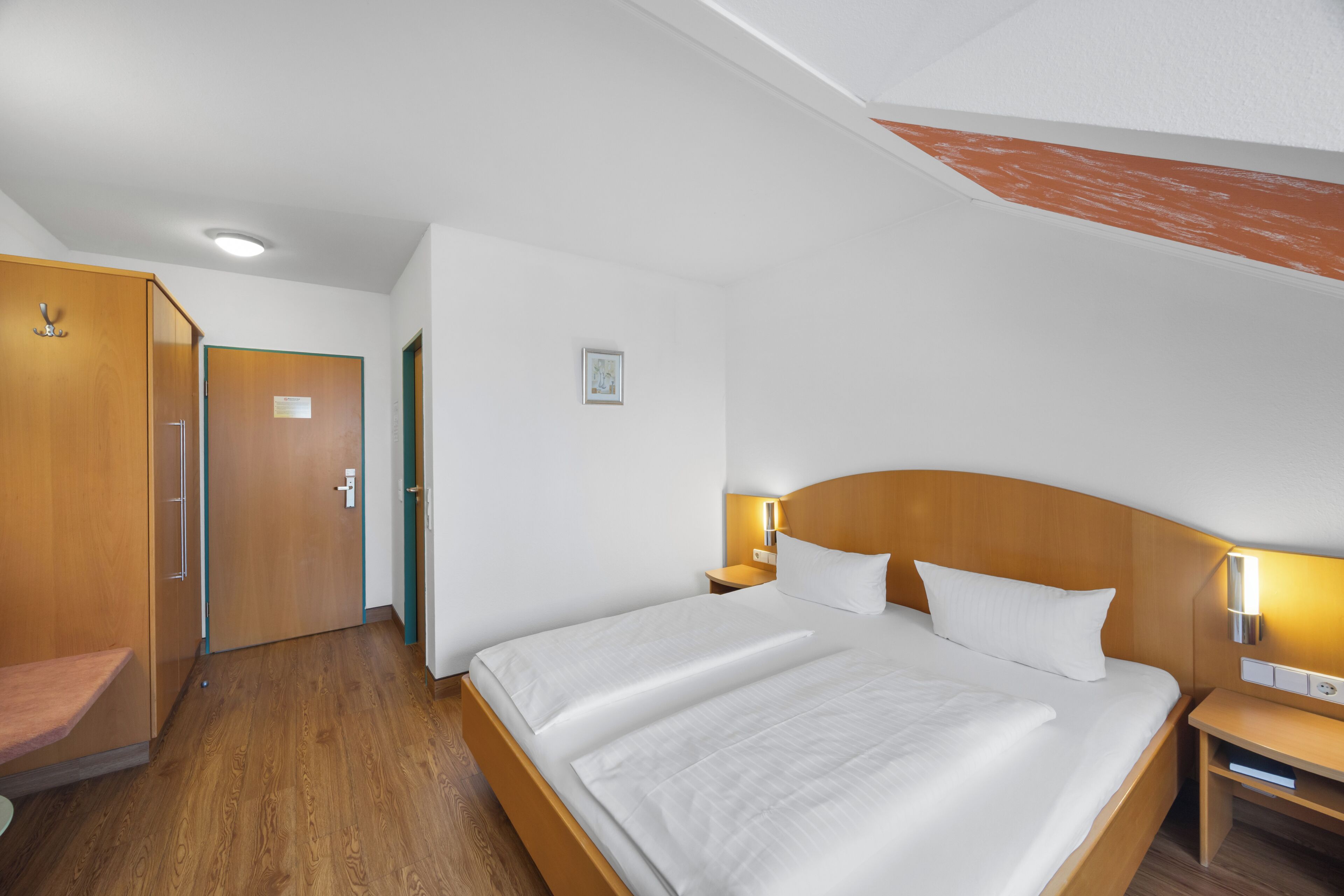 Quarto de hotel com uma cama grande, cabeceira de madeira, mesas de cabeceira com luminárias, guarda-roupa de madeira e uma pequena escrivaninha.