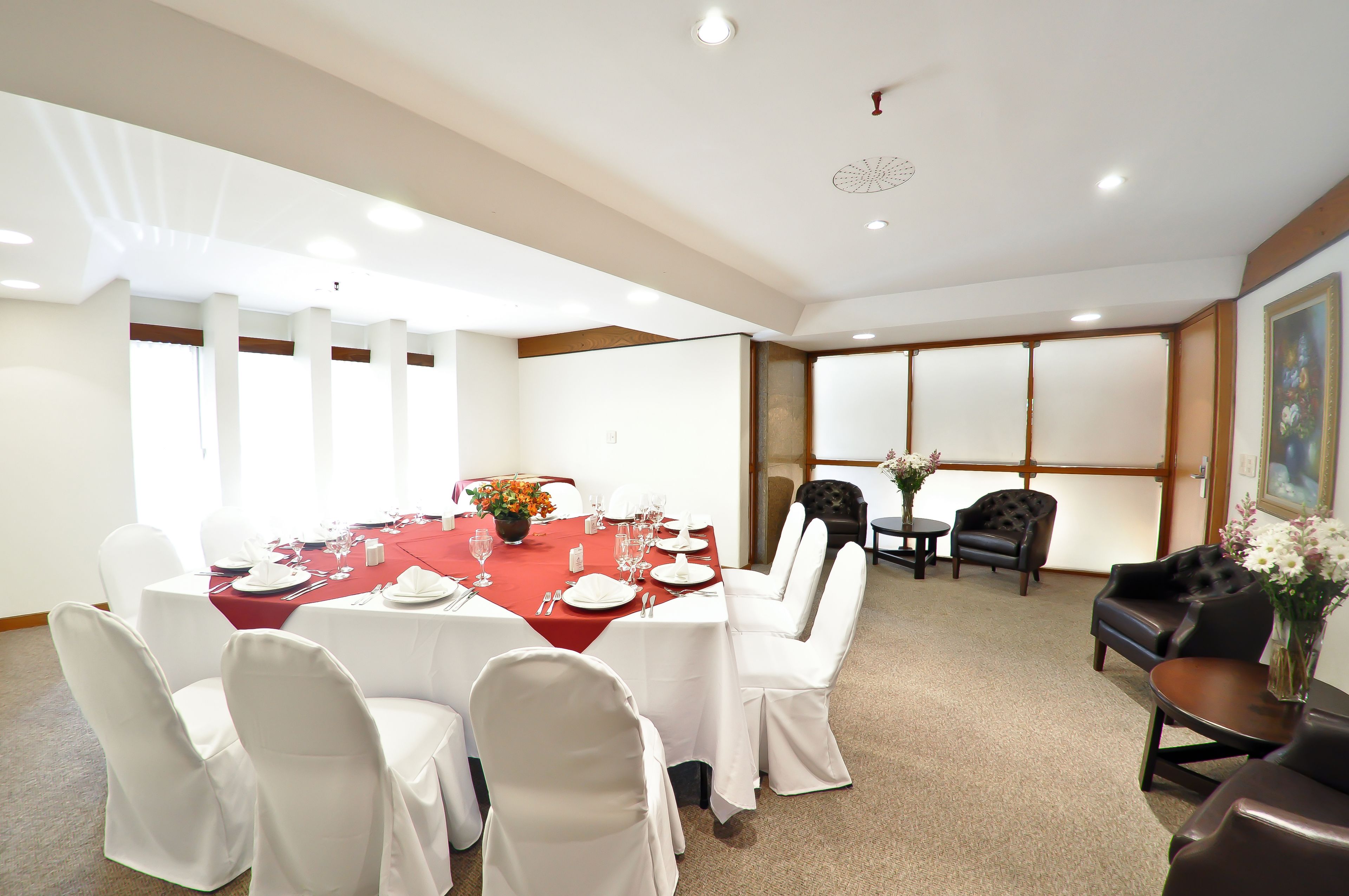 Sala de conferências com uma grande mesa posta para uma reunião, cercada por cadeiras brancas, e uma área de estar com sofás e uma pequena mesa.
